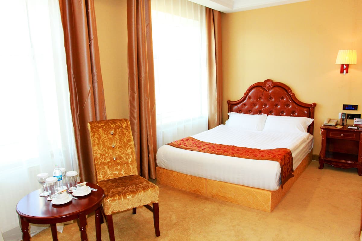 Quarto de hotel com uma cama grande, uma mesa de cabeceira de madeira, uma cadeira e uma pequena mesa com uma garrafa e copos.