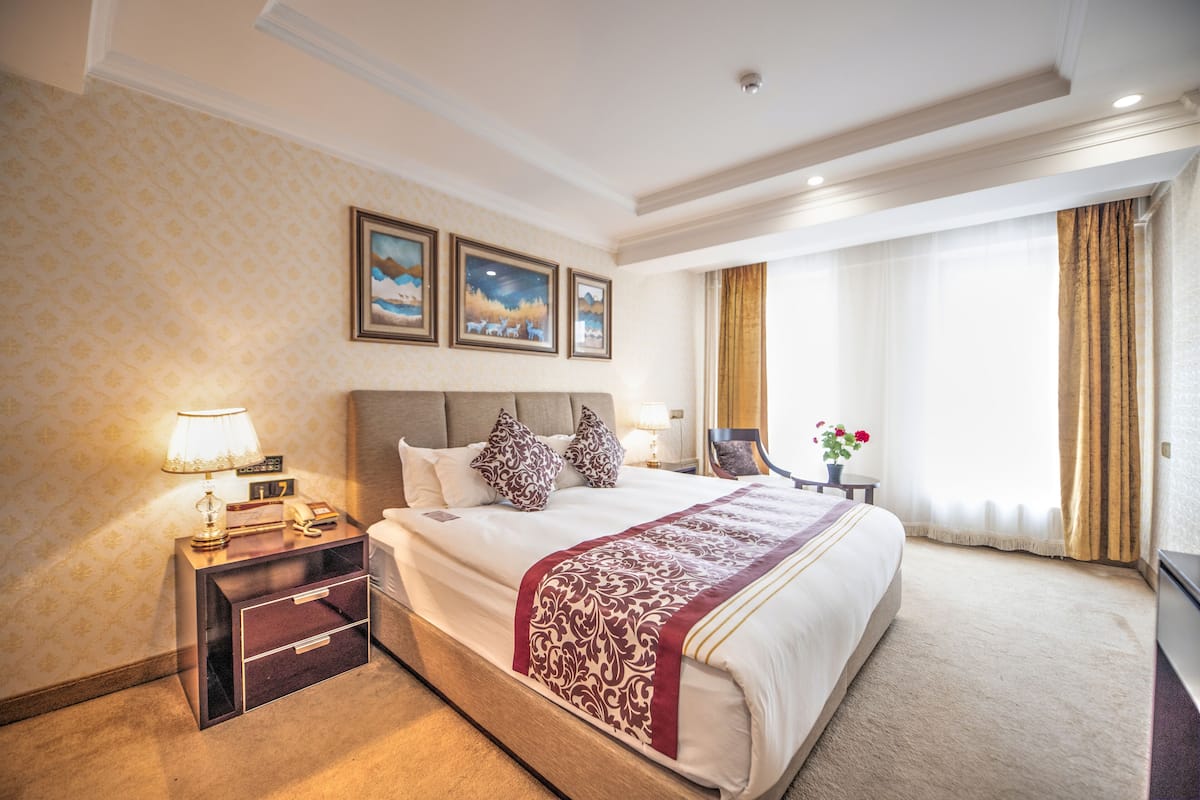 Quarto de hotel com cama grande, mesa de cabeceira com abajur, pequena mesa com vaso de flores e quadros nas paredes.