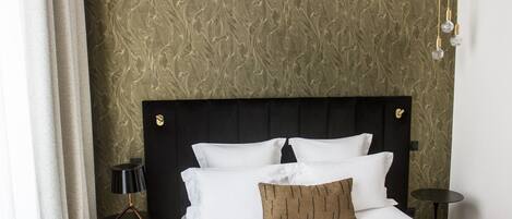 Uma cama bem arrumada com lençóis brancos e um travesseiro marrom decorativo, uma cabeceira preta e uma pequena mesa de cabeceira com uma luminária.