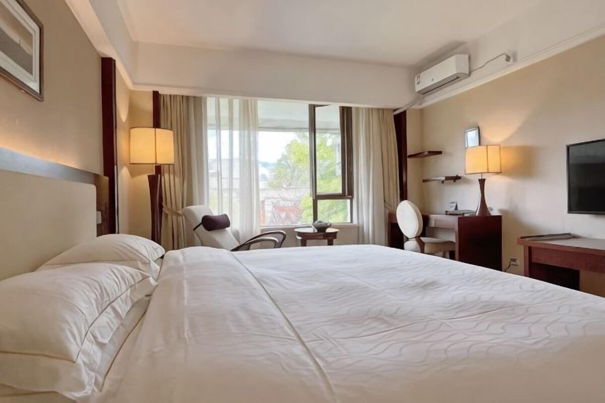 Quarto de hotel com uma cama grande, uma escrivaninha, uma cadeira, uma televisão e uma janela com cortinas.