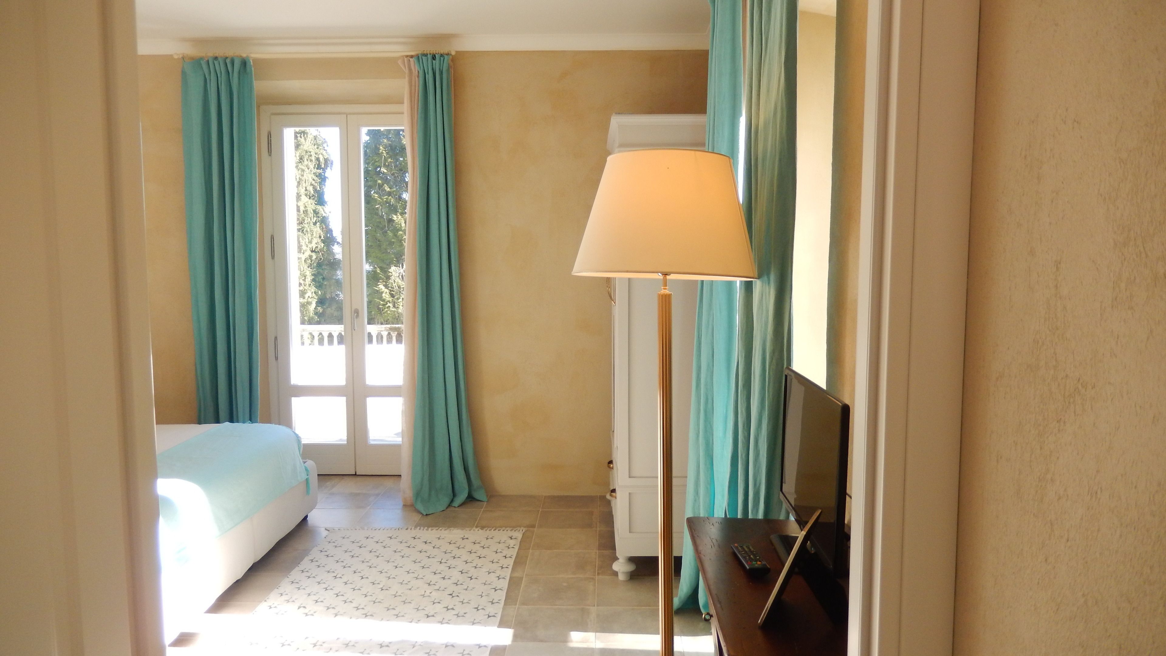 Quarto de hotel com uma cama, uma escrivaninha com abajur, uma televisão e vista para árvores pela janela.