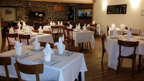 Restaurante com várias mesas dispostas, cada uma com toalha branca, guardanapo dobrado e talheres. O espaço apresenta uma parede de pedra e uma lareira, com cozinha visível ao fundo.