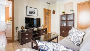 Uma sala de estar moderna com uma televisão de tela plana, uma mesa de centro de vidro, um sofá com almofadas estampadas, um armário de madeira com portas de vidro e um quadro na parede.