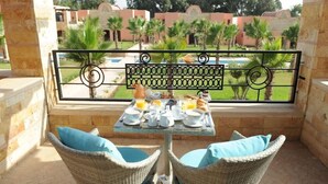 Varanda com cadeiras de vime, uma mesa com itens de café da manhã e vista para uma piscina e edifícios.