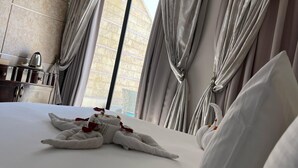 Quarto de hotel com cama bem arrumada, banheiro com chuveiro e vista para o exterior através de grandes janelas.