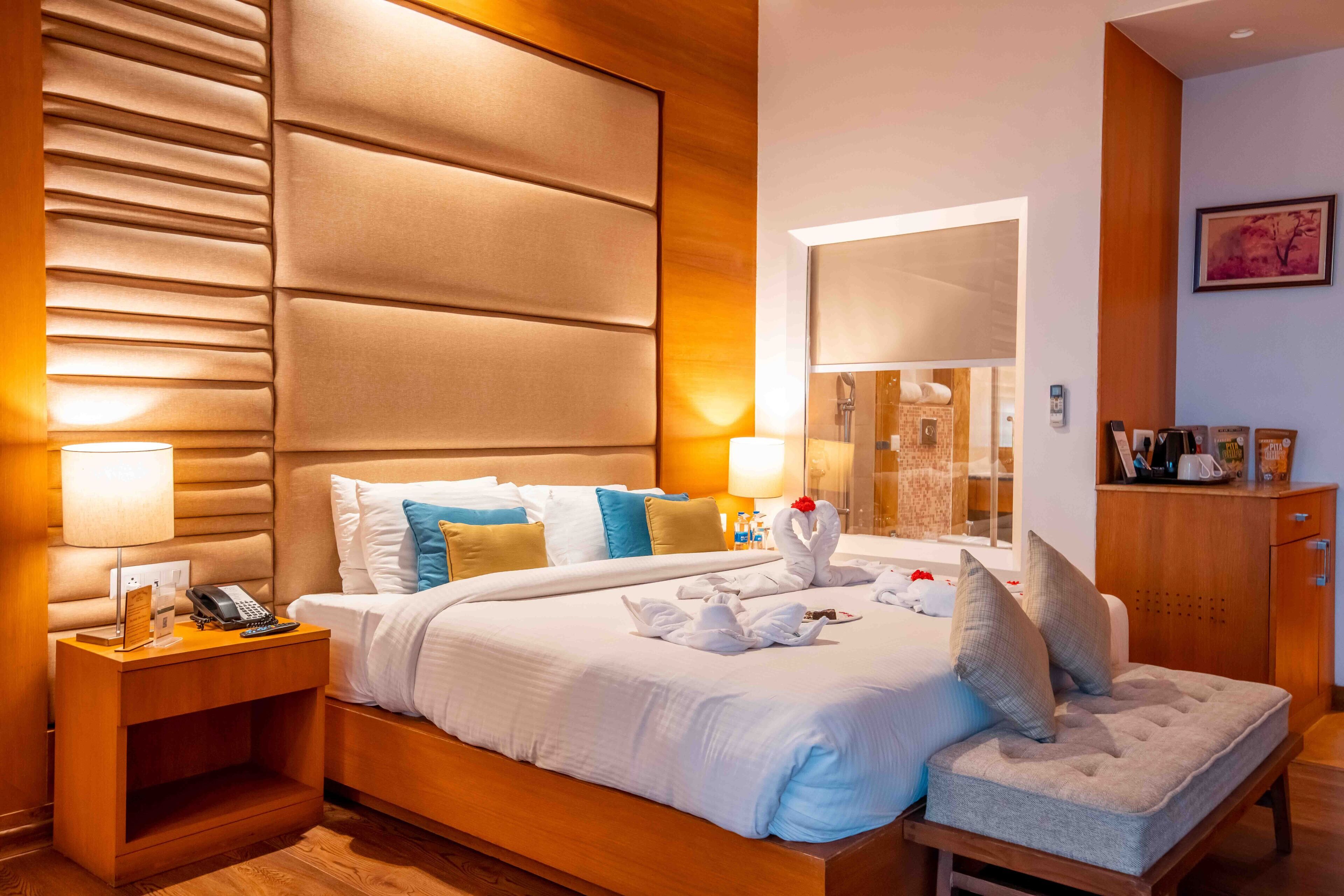 Quarto de hotel com uma cama grande, cabeceira de madeira, luminárias de cabeceira, mesa de cabeceira, guarda-roupa de madeira e um espelho.