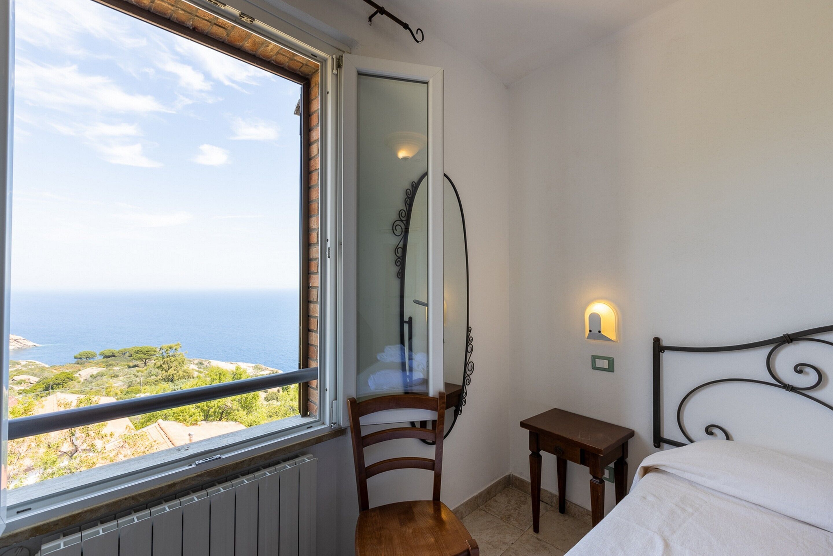 Quarto com vista para o mar, uma cadeira de madeira, uma mesa de cabeceira pequena e uma cama com cabeceira.