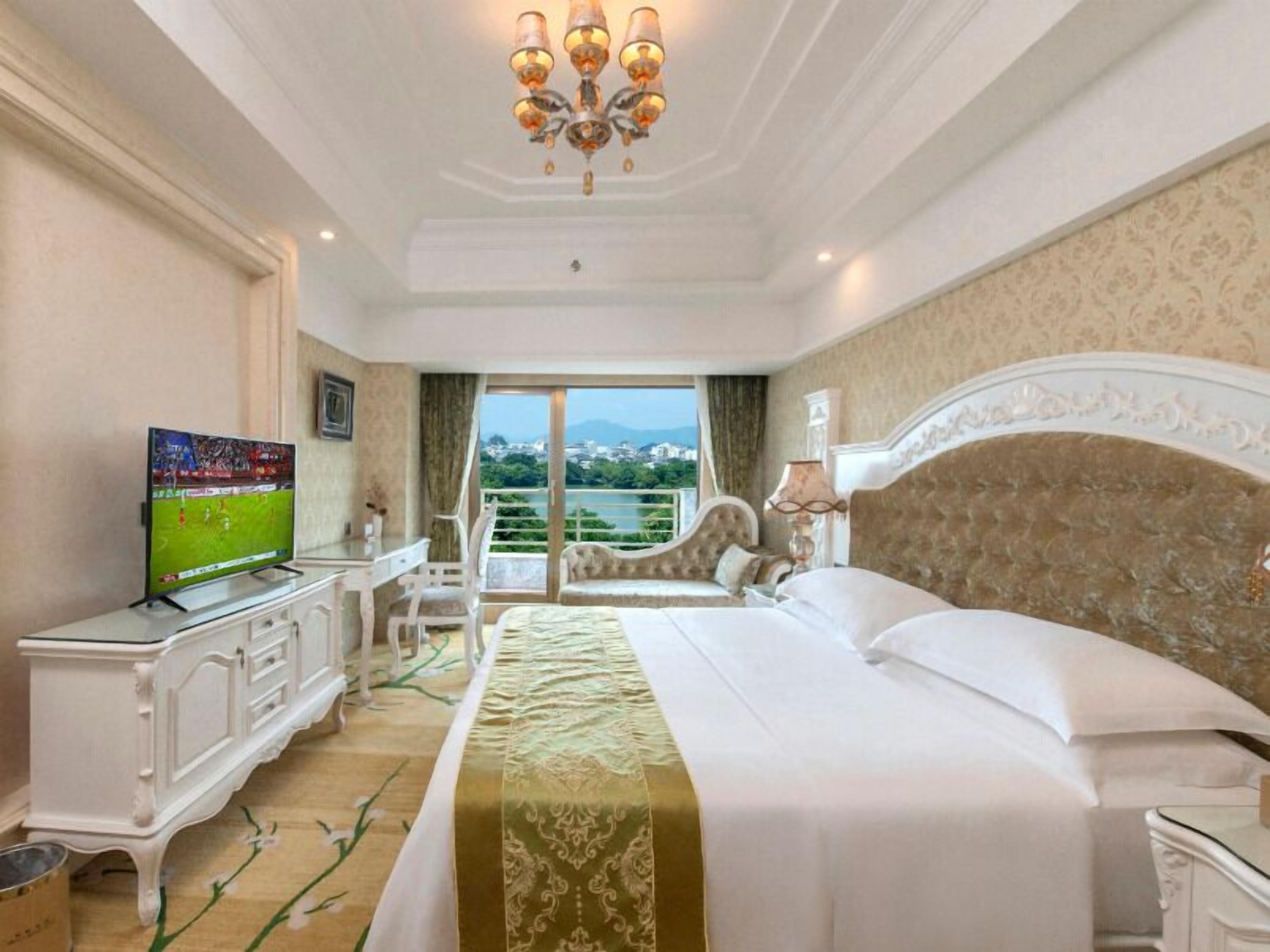 Quarto de hotel com uma cama grande, uma escrivaninha com televisão, uma cadeira e vista para o exterior.
