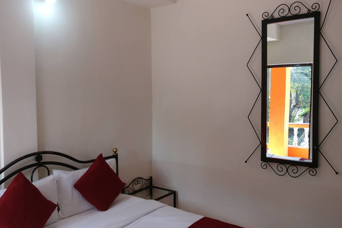 Uma cama com almofadas vermelhas e brancas, uma cabeceira de metal preto e um espelho fixado na parede refletindo uma varanda com grades laranja.