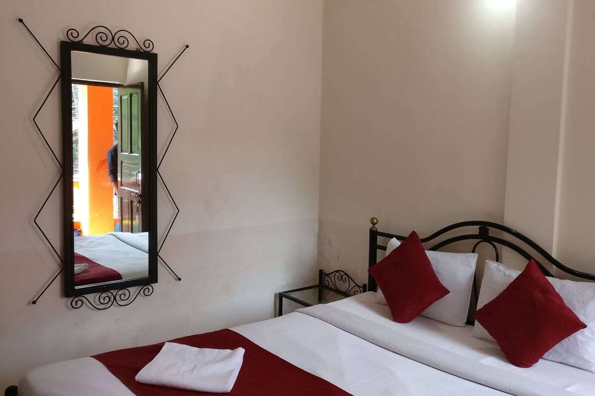 Cama bem arrumada com roupa de cama branca e vermelha, cabeceira de metal preta e espelho fixado na parede com moldura em zigue-zague.