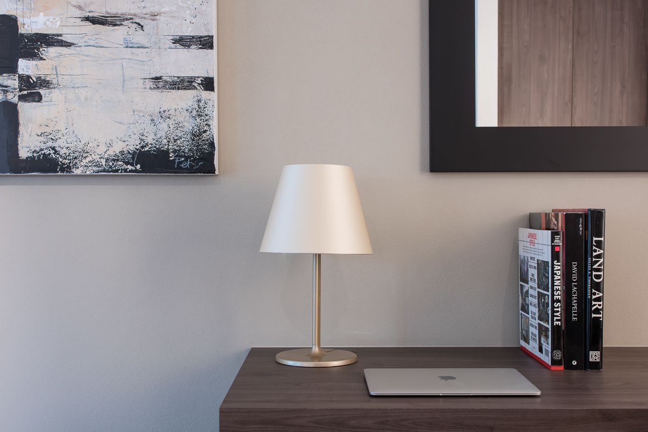 Uma escrivaninha moderna com um laptop, uma luminária branca e uma estante com livros.