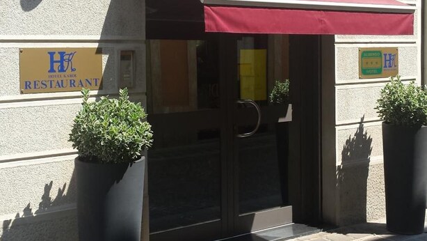 Entrada de um hotel com uma placa indicando "HOTEL KAROLE" e um toldo vermelho. Há duas plantas em vasos na frente da entrada.