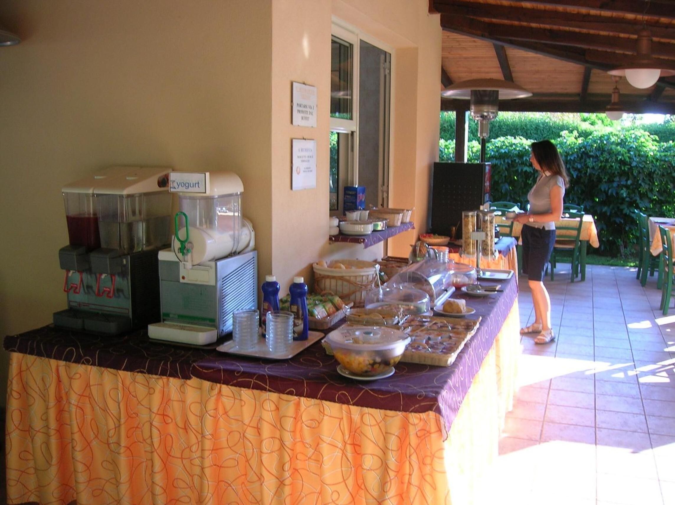 Buffet montado com dispensador de iogurte, diversos alimentos e uma pessoa ao lado do balcão.