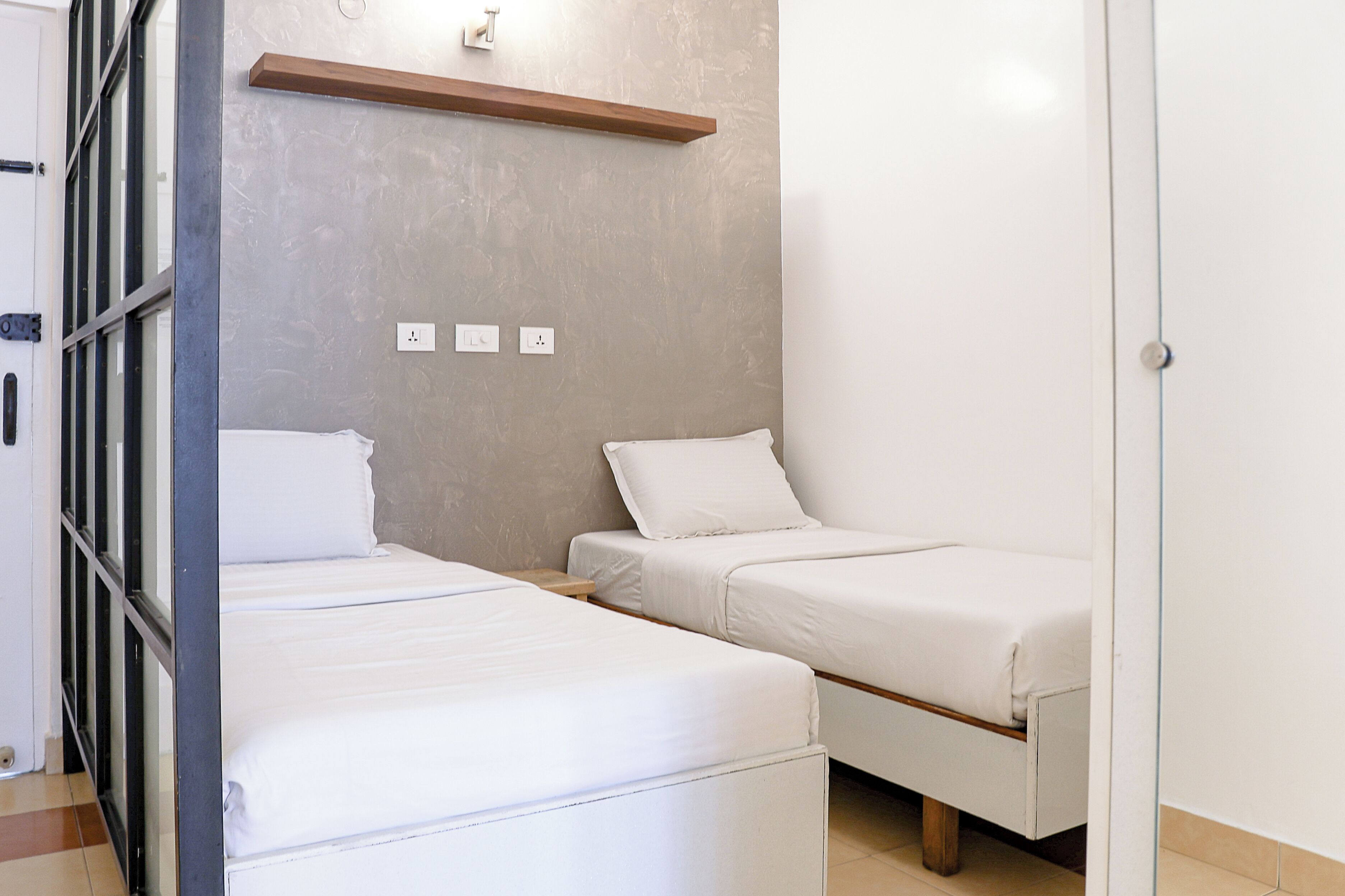 Quarto com duas camas, uma prateleira de madeira e uma parede branca com tomadas.