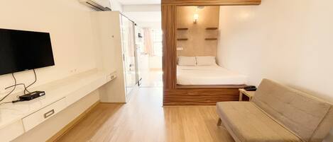 Quarto de hotel compacto com cama, sofá, televisão fixada na parede e um painel decorativo de madeira.