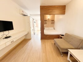 Quarto de hotel compacto com cama, sofá, televisão fixada na parede e um painel decorativo de madeira.
