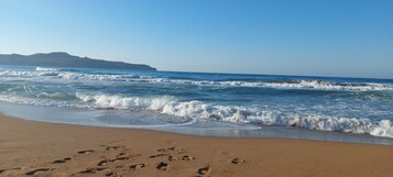 Uma praia com pegadas que levam à água, ondas quebrando na costa e uma montanha distante sob um céu claro.