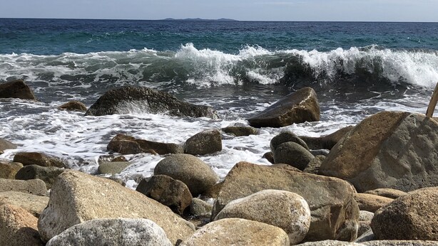 Uma linha costeira rochosa com ondas quebrando nas pedras e o oceano ao fundo.