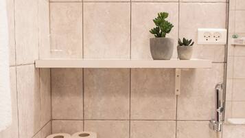 Casa de banho com azulejos bege, uma prateleira com duas plantas em vasos, um porta-papel higiénico e um painel de controlo do chuveiro.