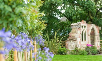 Um jardim com uma estrutura de pedra, possivelmente uma capela, rodeado por vegetação exuberante e flores roxas.