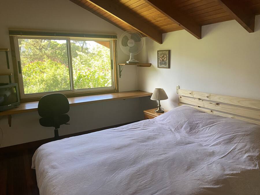 Quarto com cama, escrivaninha com cadeira, janela com vista para o verde, luminária de cabeceira e ventoinha fixada na parede.