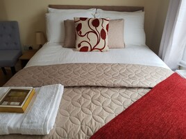 Uma cama bem arrumada com um edredom bege, um travesseiro estampado em vermelho e branco, e um cobertor vermelho.