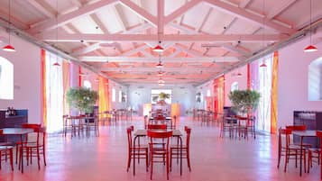Uma sala de jantar ampla com cadeiras e mesas vermelhas, janelas grandes e luminárias pendentes vermelhas.