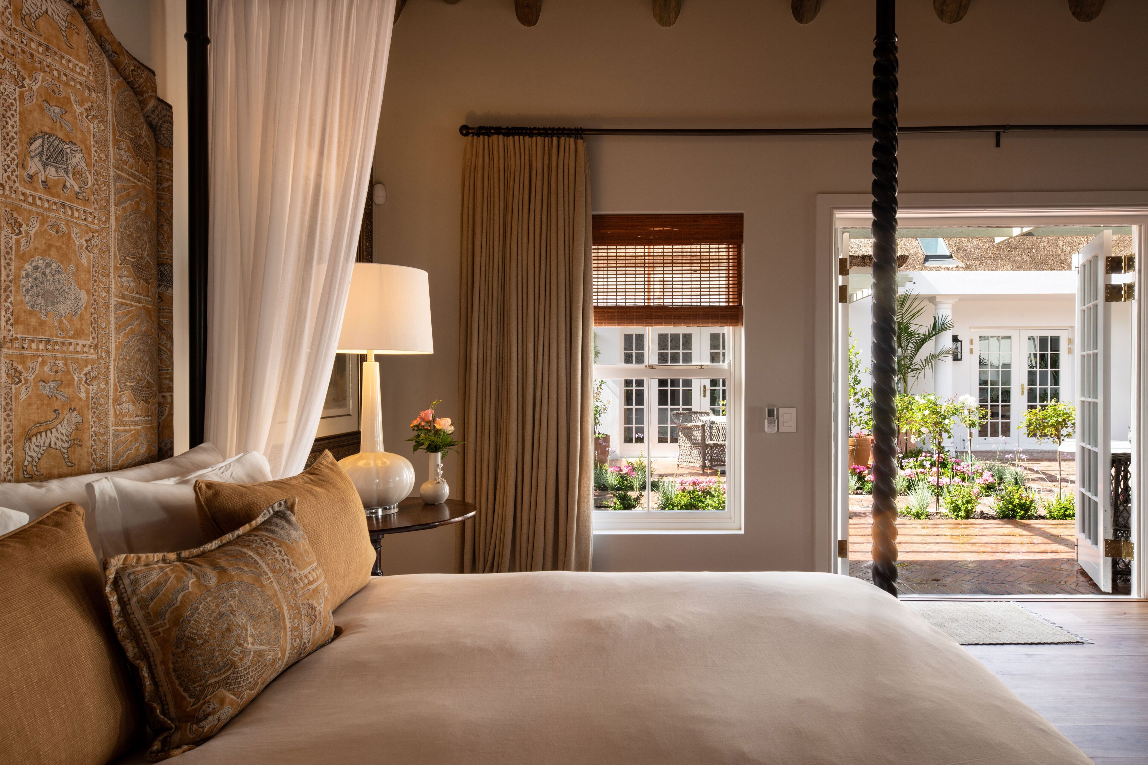 Quarto com uma cama grande, mesa de cabeceira com abajur e vista do jardim por uma porta aberta.