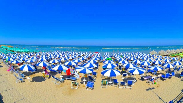 Uma praia com vários guarda-sóis azuis e brancos, espreguiçadeiras e cadeiras de praia.