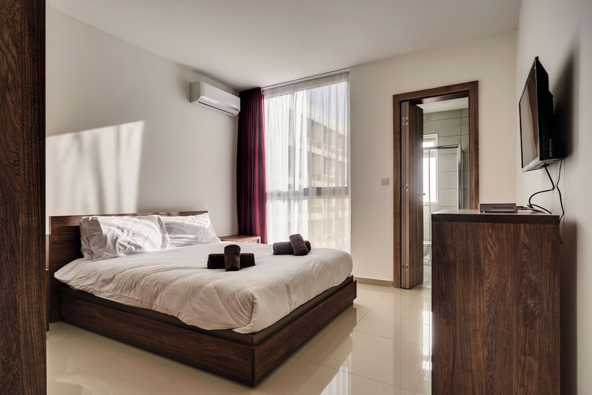 Quarto de hotel moderno com uma cama grande, cabeceira de madeira, TV fixada na parede e uma janela ampla com cortinas leves.