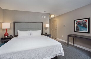 Quarto de hotel com uma cama grande, duas mesas de cabeceira, um criado-mudo, uma escrivaninha, uma cadeira, uma porta e um quadro na parede.