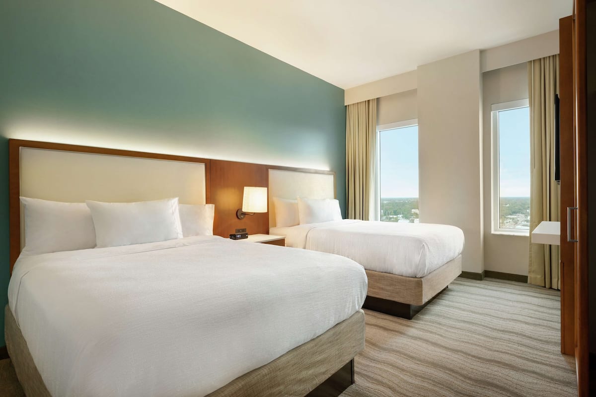 Quarto de hotel com duas camas, mesa de cabeceira, luminária, janela com cortinas e vista para a cidade.