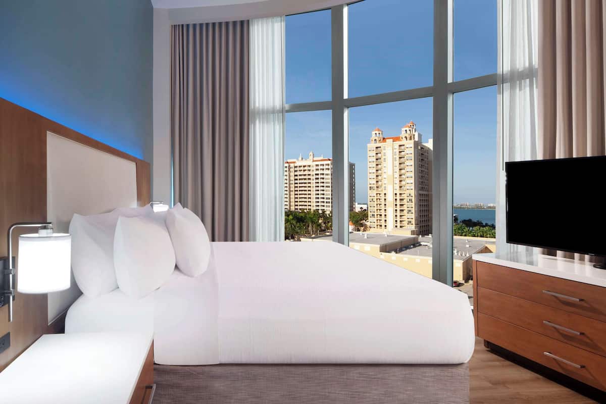 Quarto de hotel com uma cama grande, televisão, escrivaninha e vista para edifícios altos e um corpo d'água.