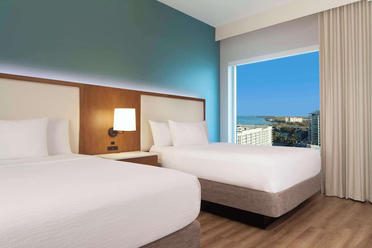 Quarto de hotel com duas camas, uma mesa de cabeceira com abajur e uma janela ampla com vista para a cidade.