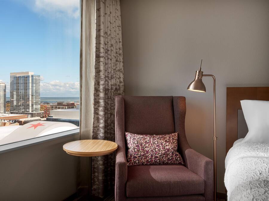 Quarto de hotel com vista para a cidade, uma mesa redonda de madeira, uma poltrona marrom com almofada florida e um abajur.
