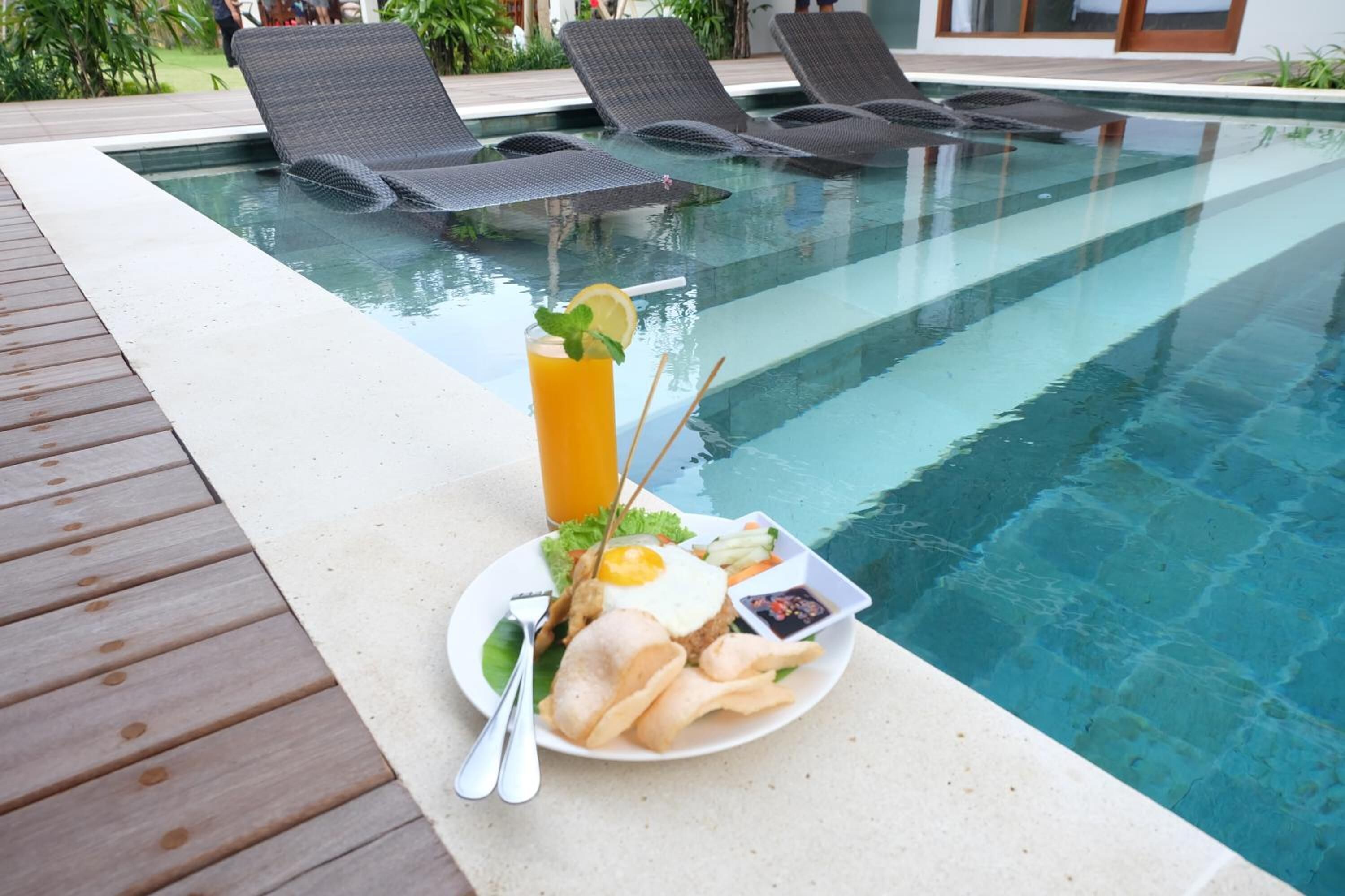 Uma área de refeições com mesa à beira da piscina, um prato com comida e um copo de suco de laranja.