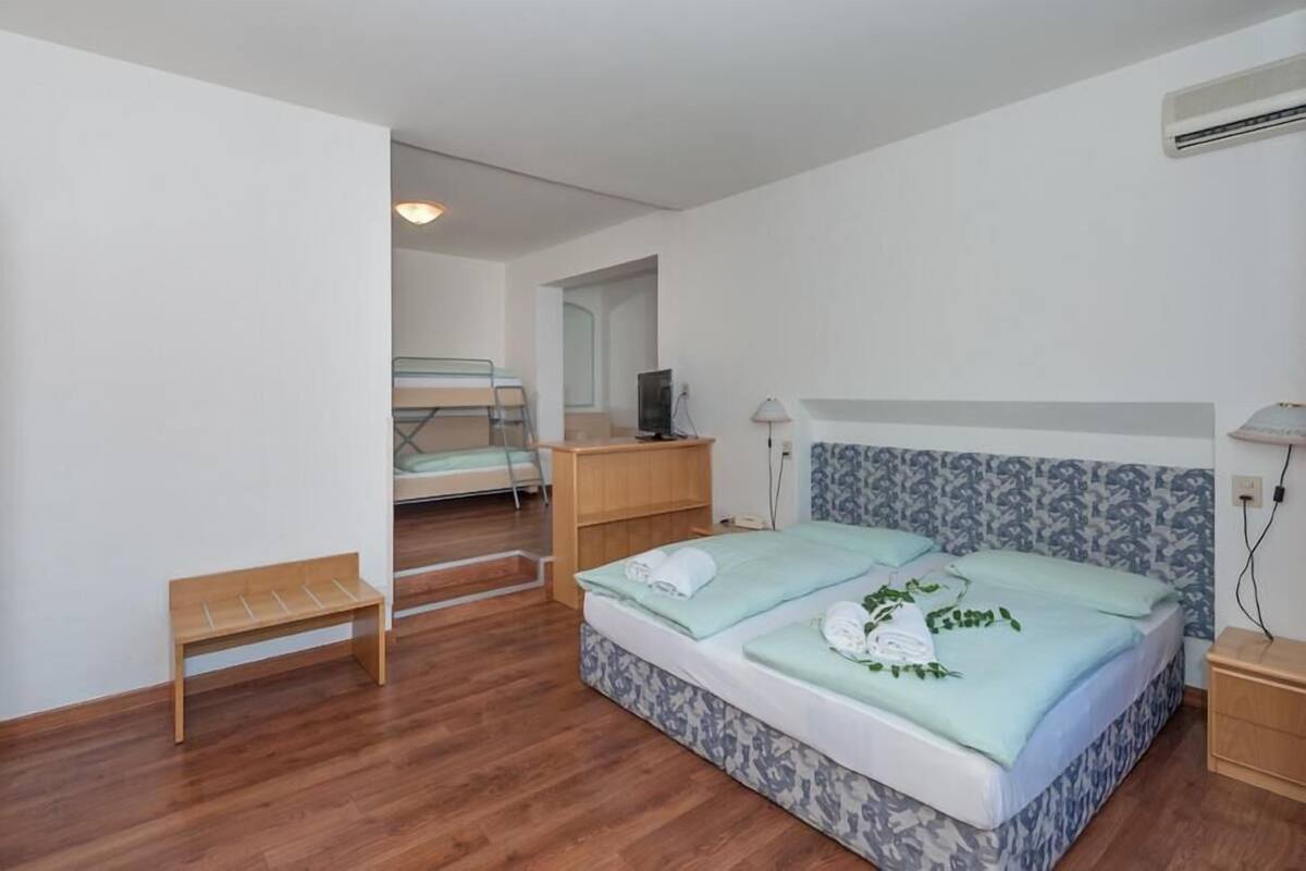 Quarto de hotel com cama, um banco, escrivaninha, televisão e aparelho de ar condicionado.