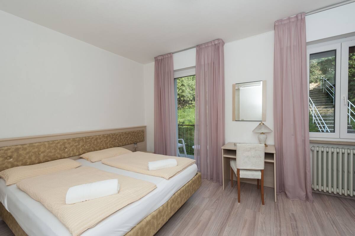 Quarto de hotel com duas camas, uma escrivaninha, uma cadeira, janela com cortinas e vista para uma escadaria.