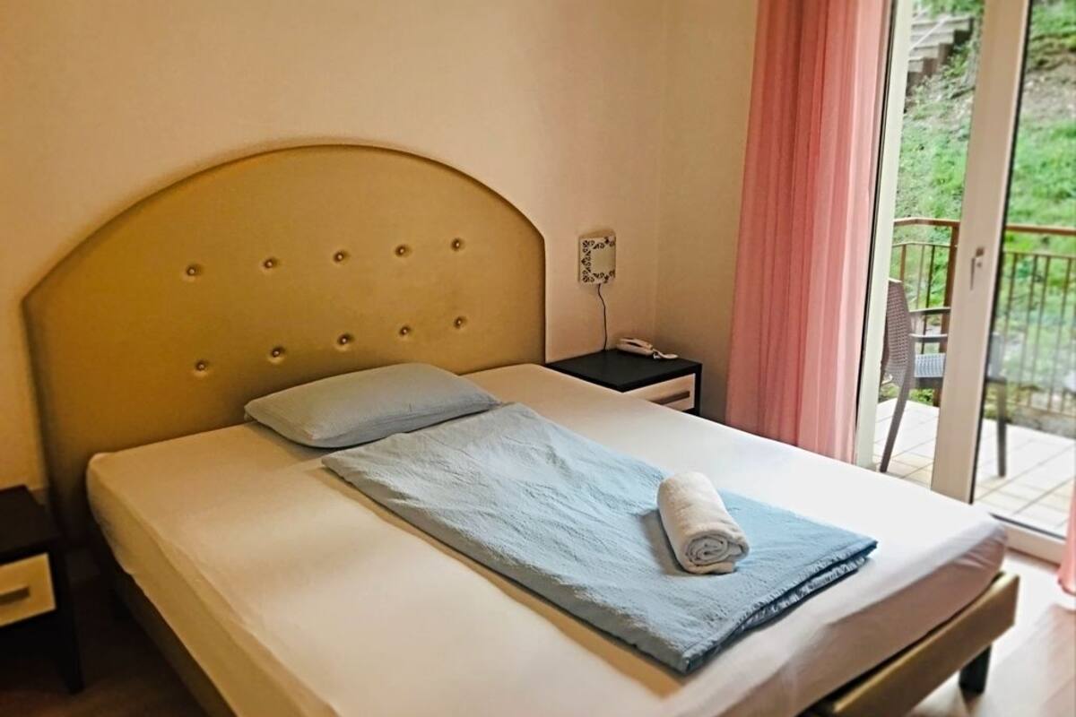 Quarto de hotel com uma cama, mesa de cabeceira, tomada e vista para uma varanda com vegetação.