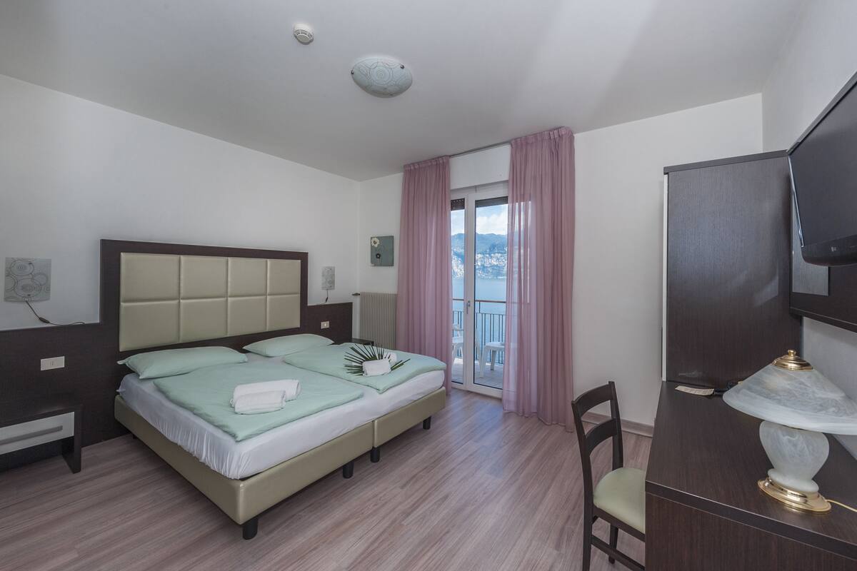 Quarto de hotel com cama, escrivaninha com luminária, cadeira e vista para o exterior.