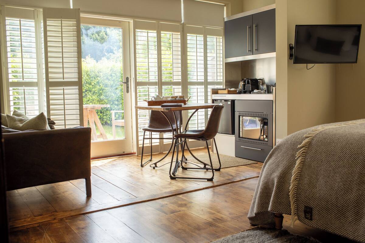 Quarto com piso de madeira, uma cama, uma mesa com cadeiras, televisão e cozinha equipada com armários e fogão.