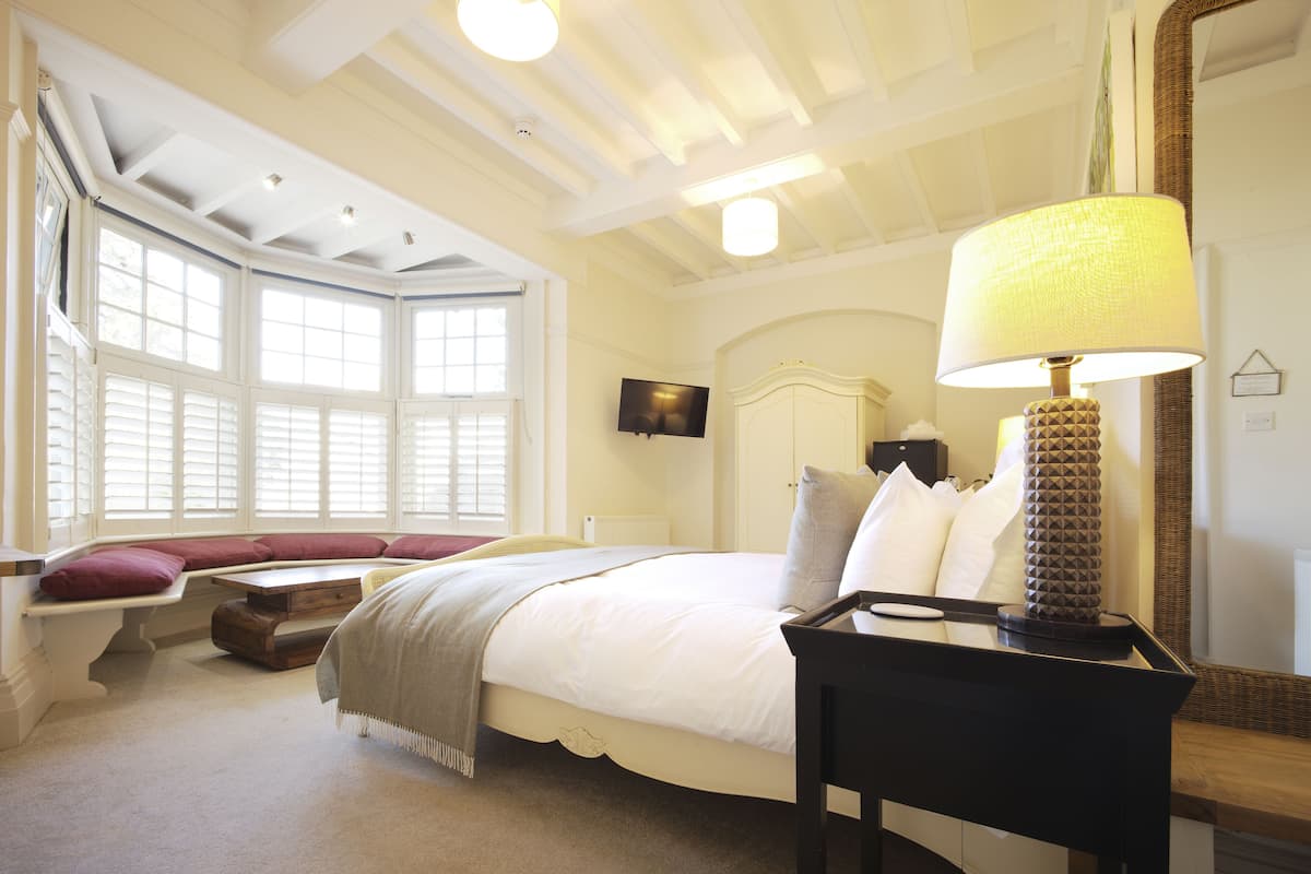 Quarto com uma cama grande, um banco junto à janela, uma mesa de cabeceira e um abajur.