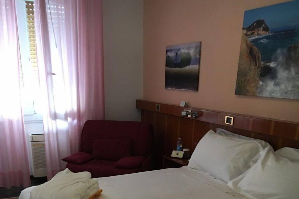 Quarto de hotel com uma cama grande, uma poltrona vermelha, um quadro na parede e uma mesa de cabeceira com abajur.