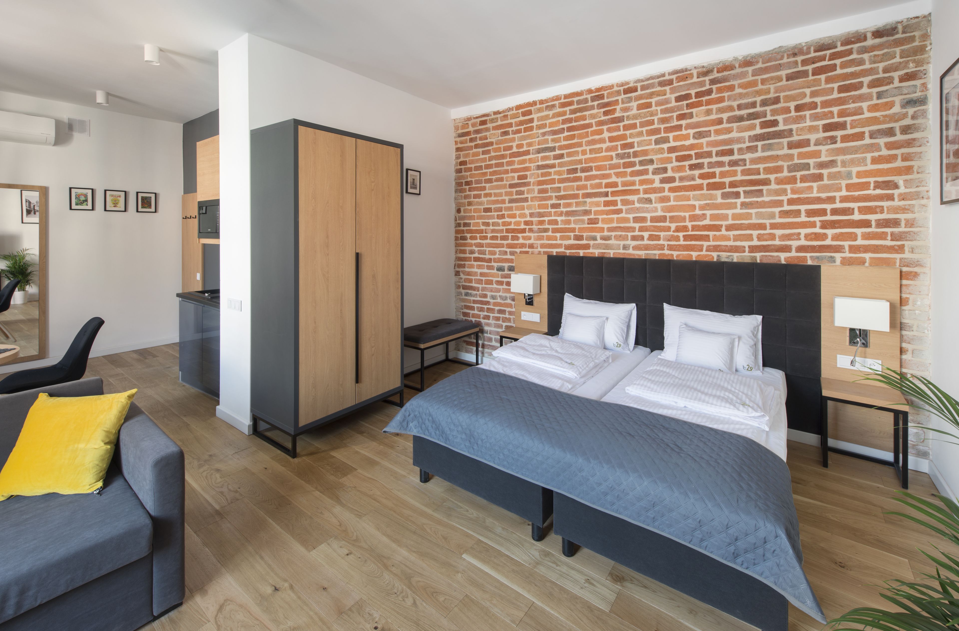 Quarto moderno com parede de tijolos, cama com roupa de cama branca, sofá cinza com almofada amarela e guarda-roupa de madeira.