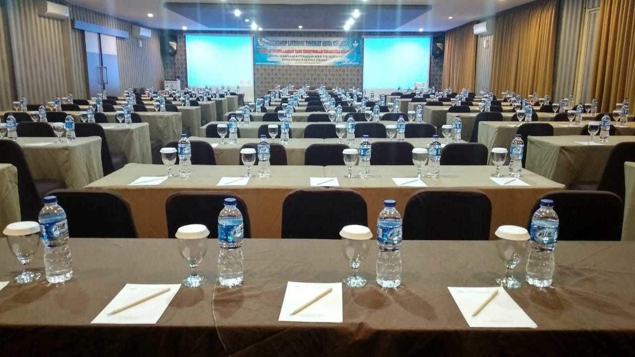 Sala de conferências com mesas alinhadas, cada uma com garrafas de água e copos, e uma tela grande na frente.