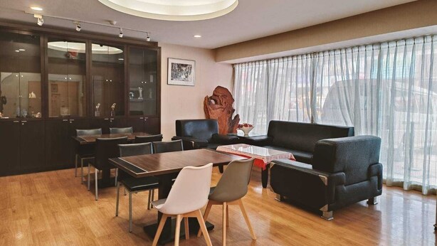 Uma sala de jantar moderna com uma mesa de madeira, cadeiras e um sofá. Há uma janela grande com cortinas leves e vista para o exterior.