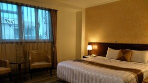 Quarto de hotel com uma cama grande, uma cadeira, uma mesa pequena e uma mesa de cabeceira com abajur.