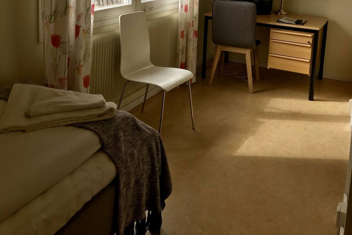 Quarto com uma cama, uma escrivaninha com cadeira, um radiador e uma janela com cortinas.