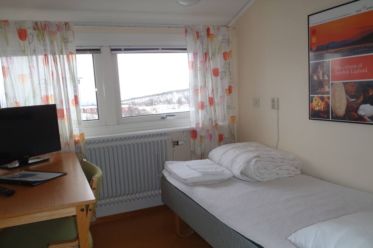 Quarto de hotel com cama, escrivaninha com computador, radiador e janela com cortinas.