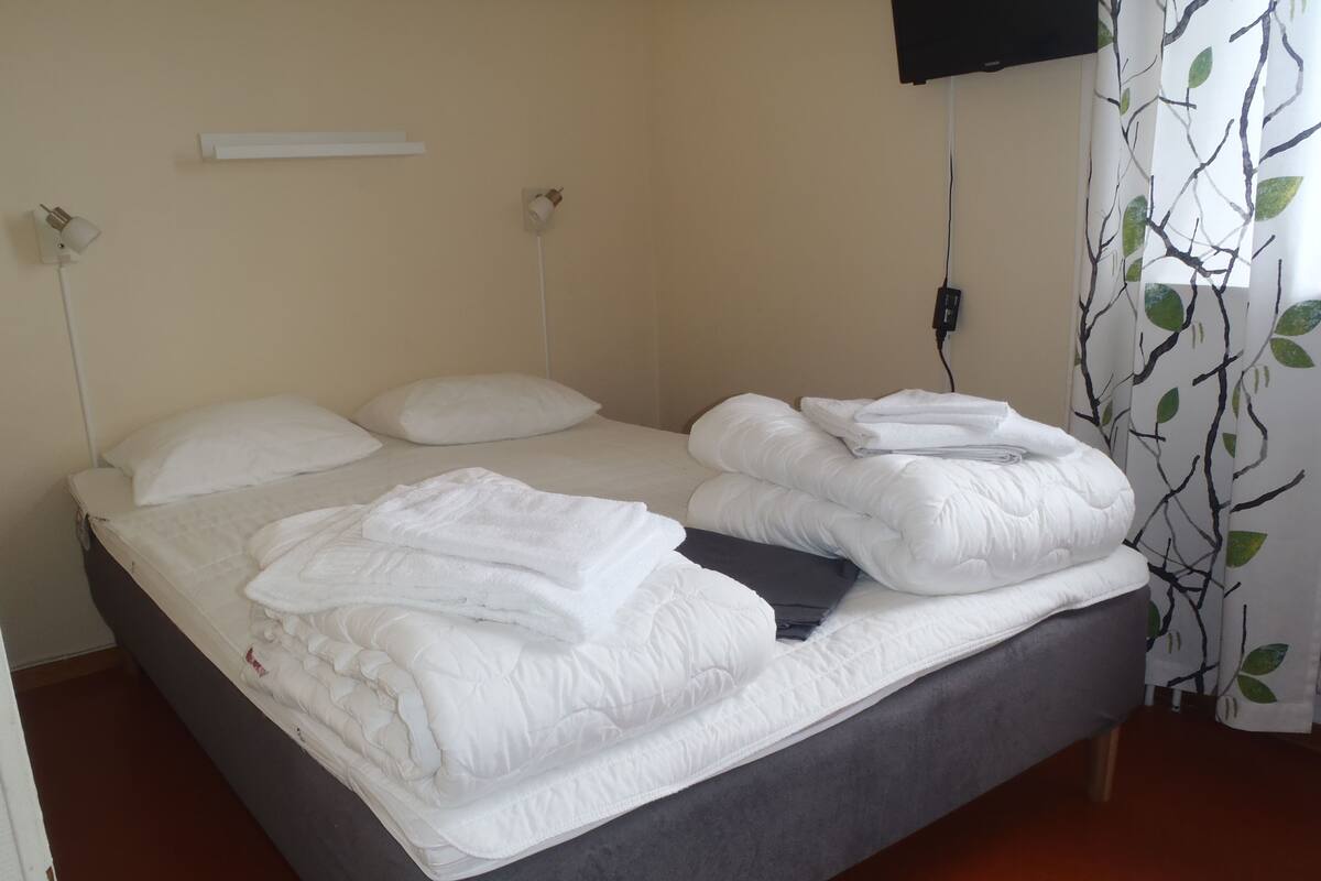 Cama bem arrumada com lençóis brancos e colchão cinza em quarto com parede lisa e cortina estampada com folhas.